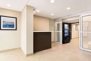 Uma área de recepção com balcão, um quadro na parede, máquina de venda automática e porta de vidro para outro espaço.