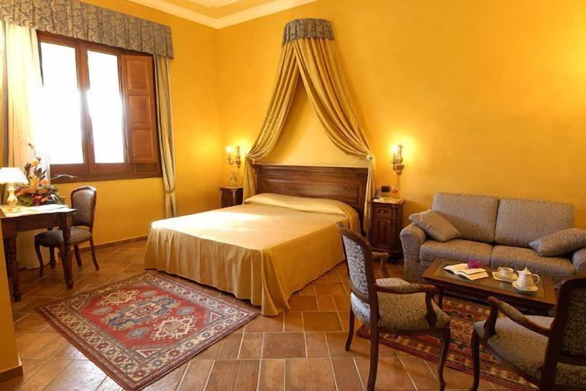 Quarto de hotel com cama, sofá, mesa de jantar com cadeiras, pequena mesa com um livro e chaleira, e tapete estampado.