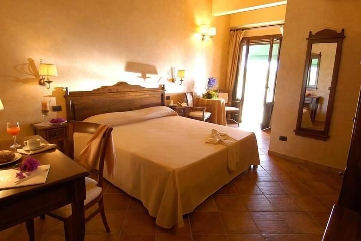 Quarto de hotel com cama de casal, escrivaninha com luminária, cadeira e mesa pequena com bebida e doces.