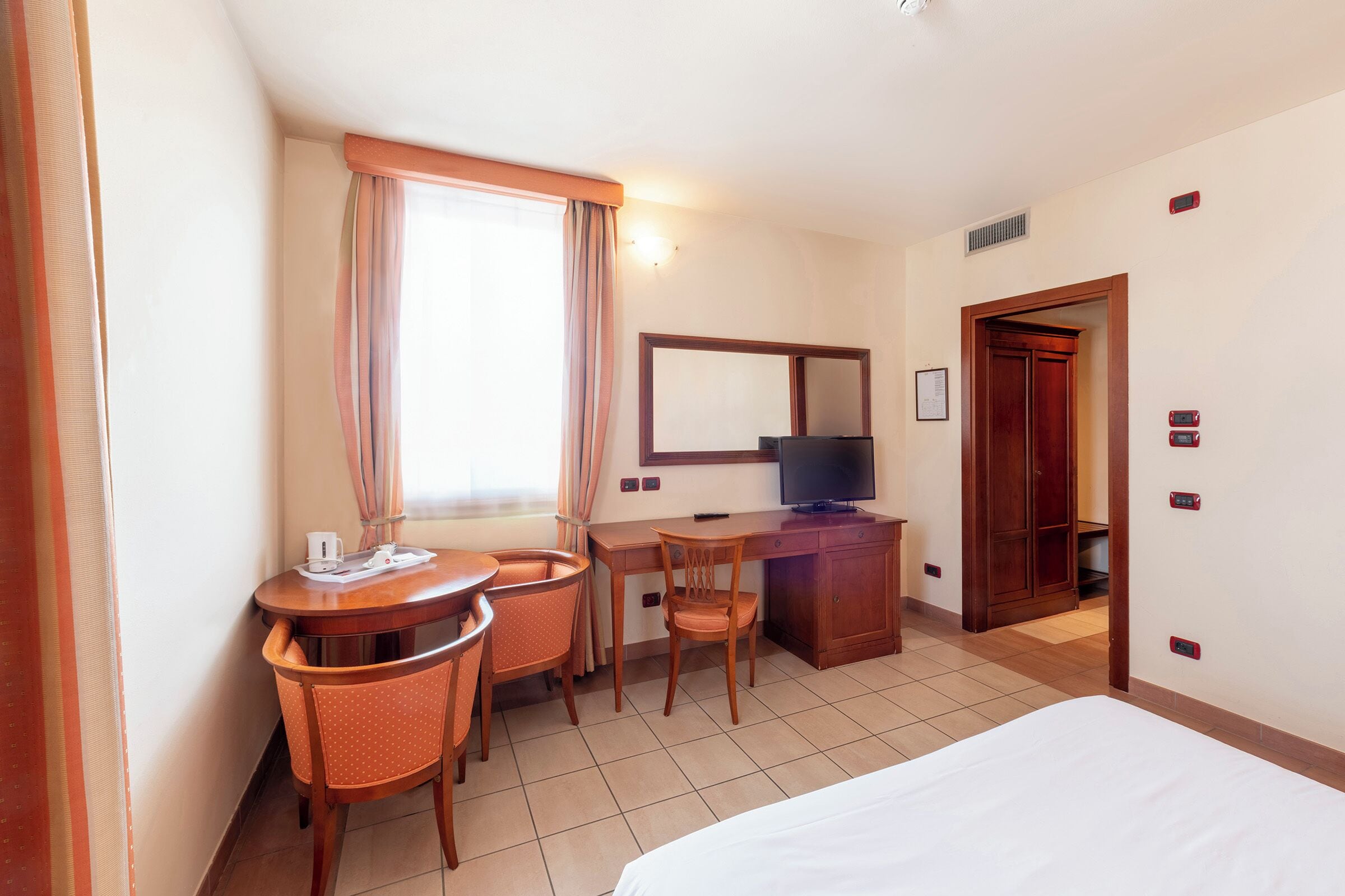 Quarto de hotel com cama, escrivaninha com cadeira, televisão e guarda-roupa.