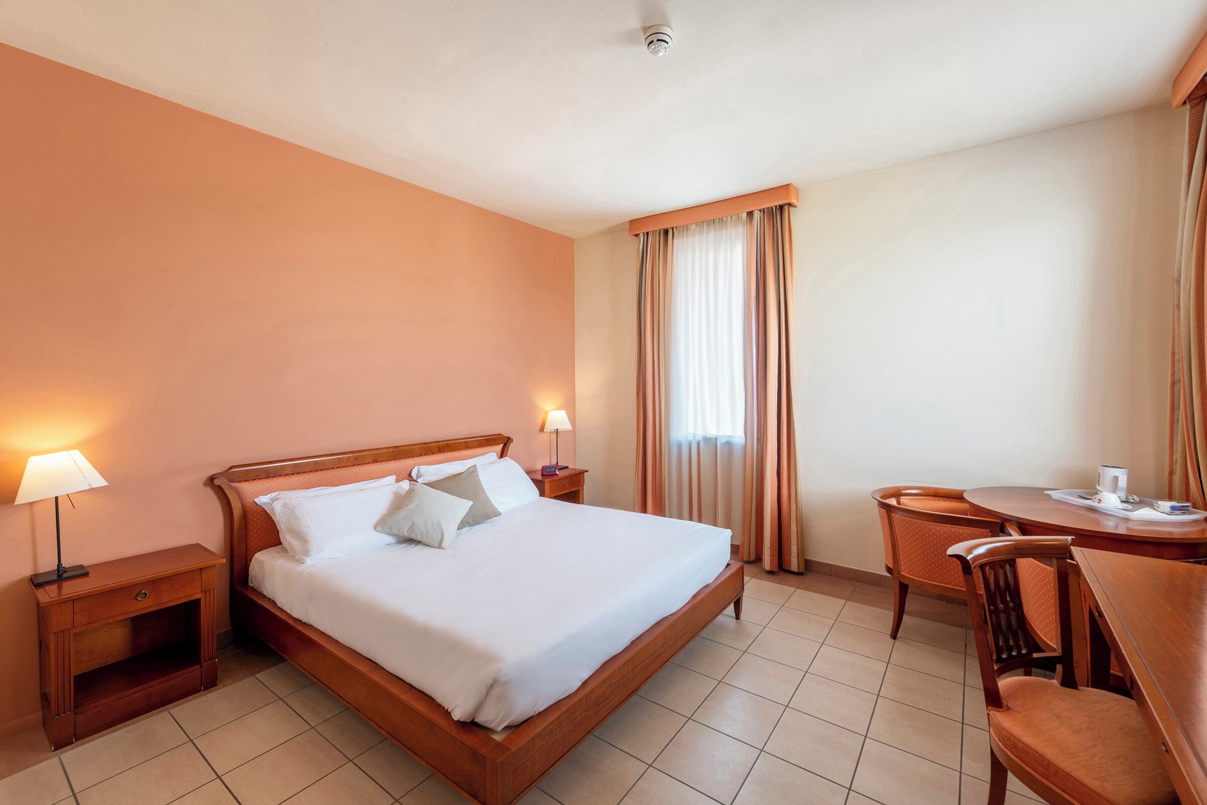 Quarto de hotel com uma cama grande, duas mesas de cabeceira com luminárias, uma pequena mesa redonda com cadeiras e uma janela com cortinas.