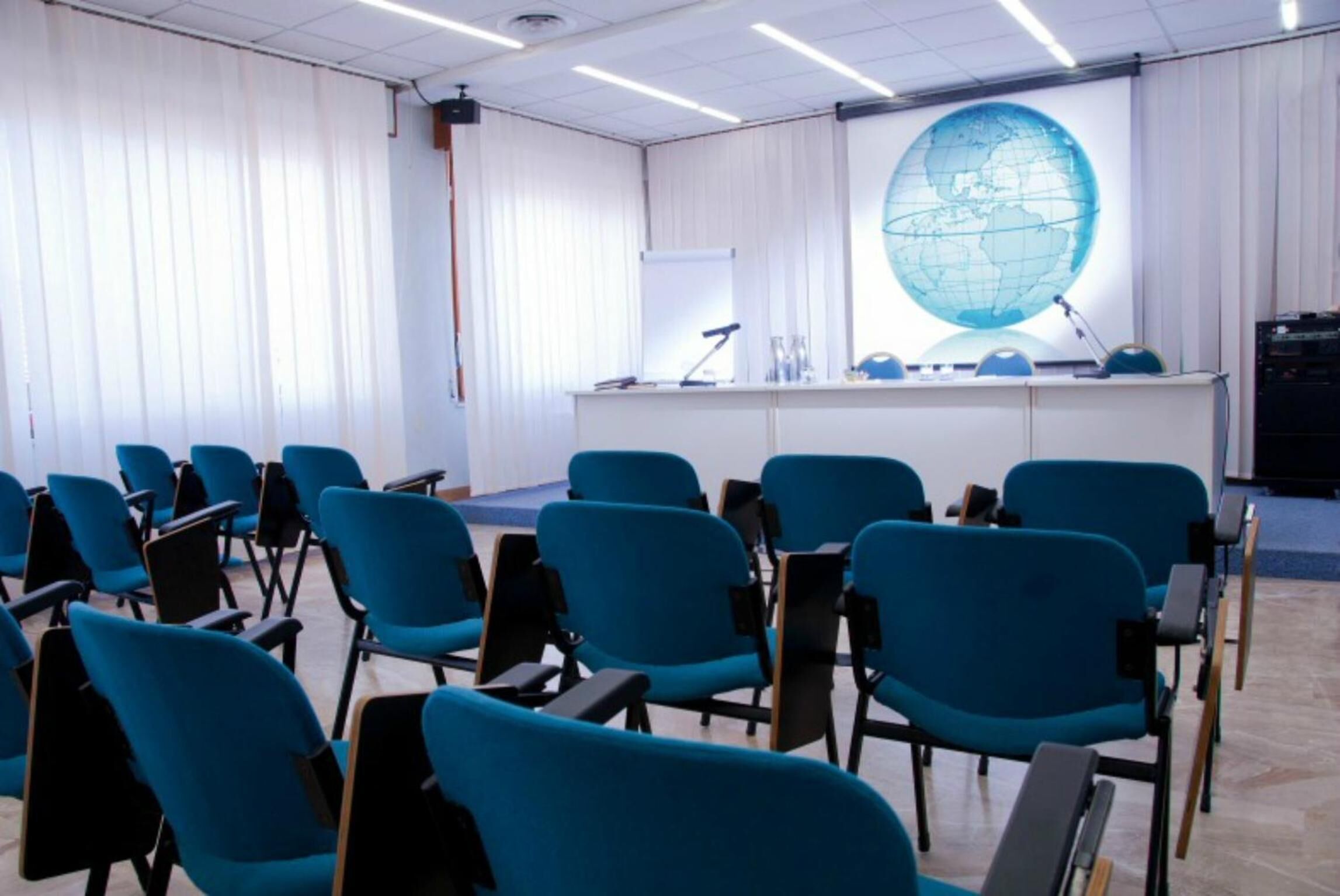 Uma sala de conferências com fileiras de cadeiras azuis, um quadro branco, projeção de um globo terrestre e um púlpito com microfone.