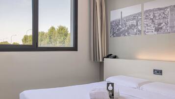 Quarto de hotel com cama, janela com vista para árvores, fotografia de uma paisagem urbana na parede e telefone na mesa de cabeceira.