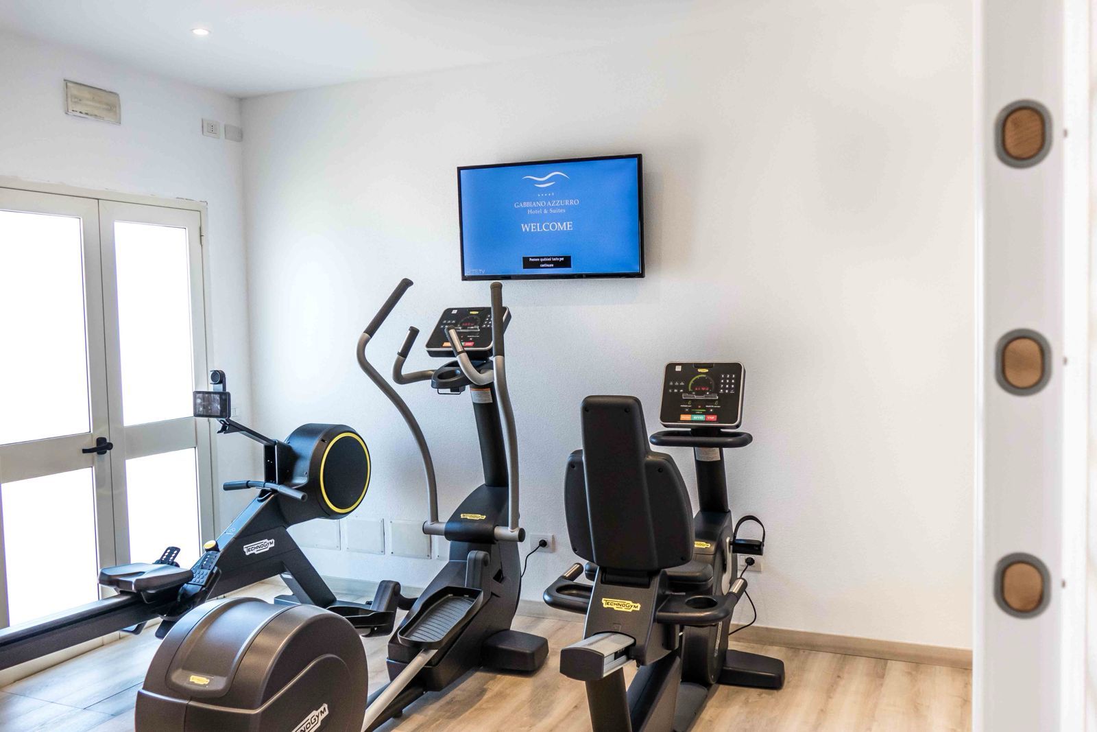 Ginásio com equipamentos de cardio, uma TV exibindo uma mensagem de boas-vindas e uma grande janela.