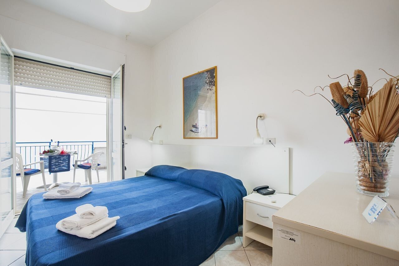 Quarto de hotel com cama, escrivaninha, cadeira, pequena mesa com um vaso e vista para a varanda.