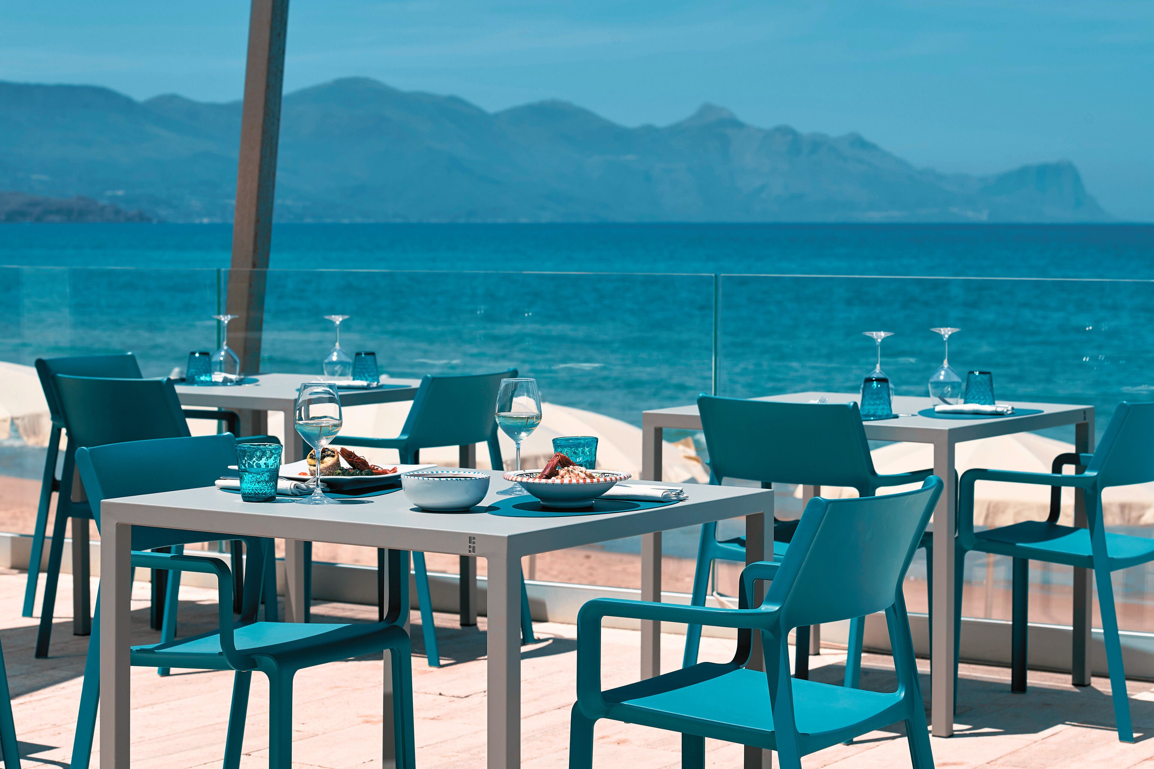 Um restaurante à beira-mar com mesas e cadeiras, vista para o oceano e montanhas ao fundo.