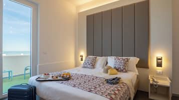 Quarto de hotel com uma cama grande, vista para o mar, bandeja de doces, mala e telefone ao lado da cama.