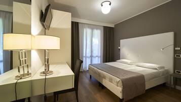Quarto de hotel com cama, escrivaninha com abajur, cadeira, espelho grande e janela com cortinas.