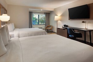 Quarto de hotel com duas camas, uma mesa, uma cadeira, uma TV e janela com vista para árvores.