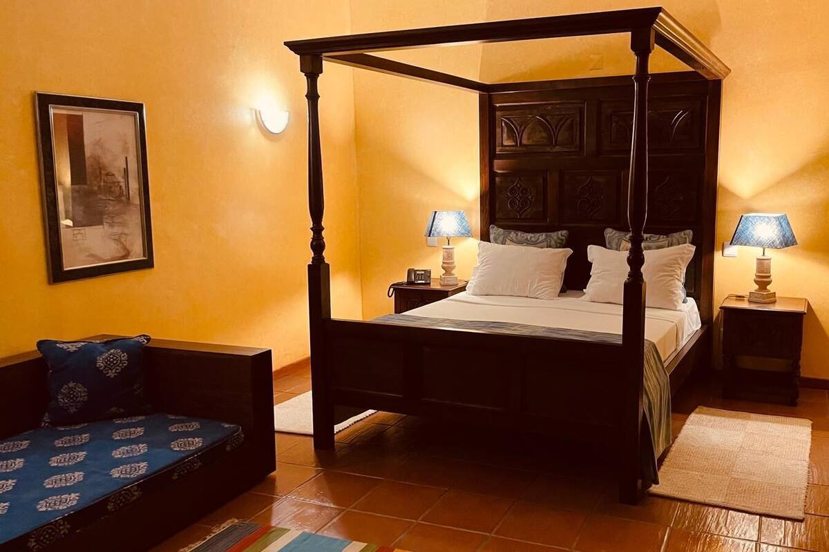 Uma cama com dossel e roupa de cama branca, duas mesas de cabeceira com abajures, um sofá azul estampado e um quadro na parede.