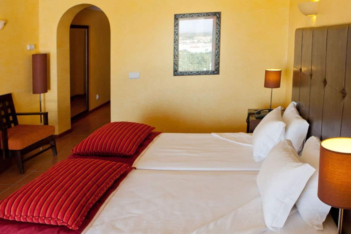 Quarto de hotel com cama, duas luminárias de cabeceira, uma cadeira e vista para o exterior pela janela.