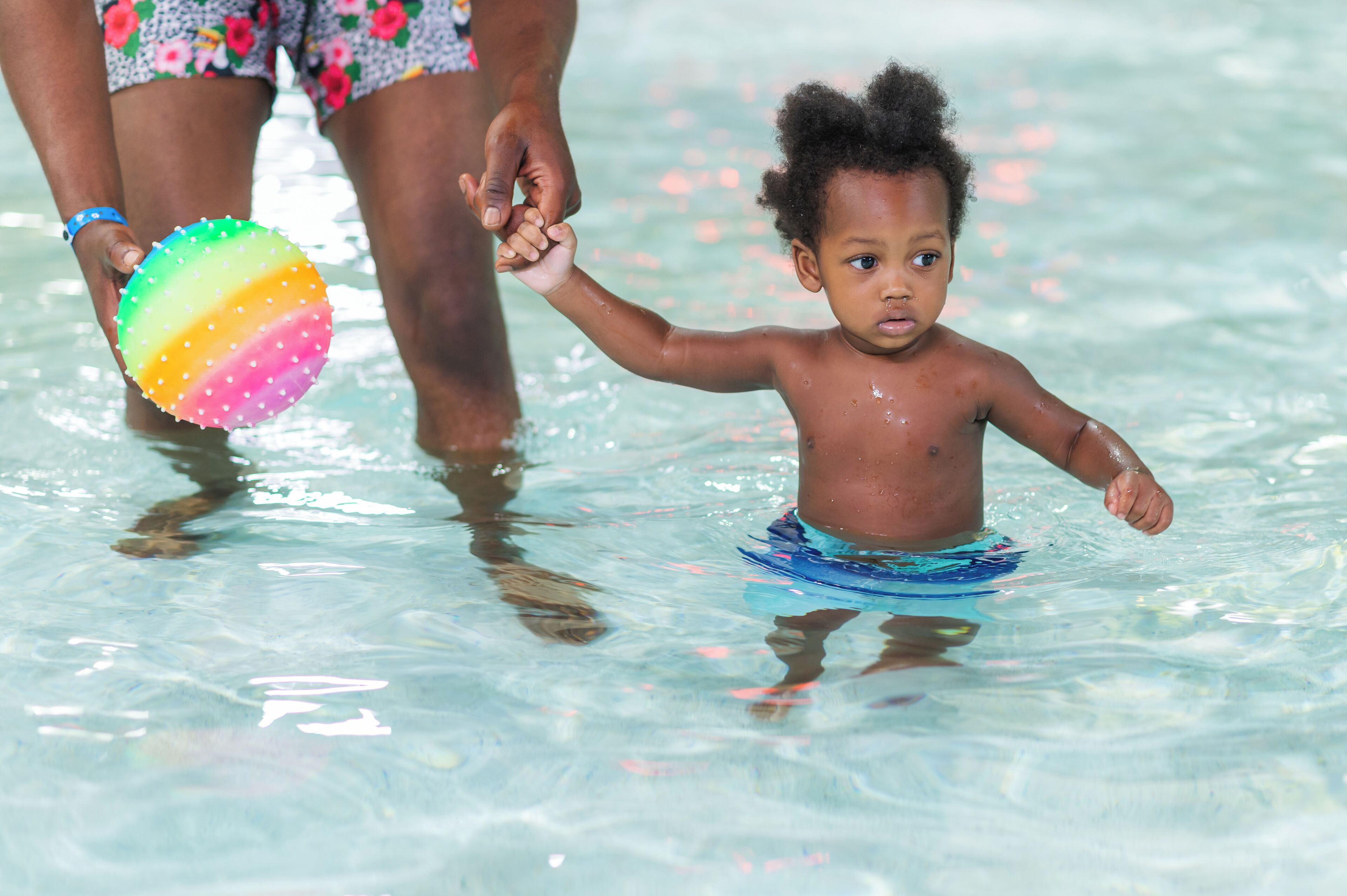 Criança em piscina sendo segurada por um adulto que segura uma bola colorida.