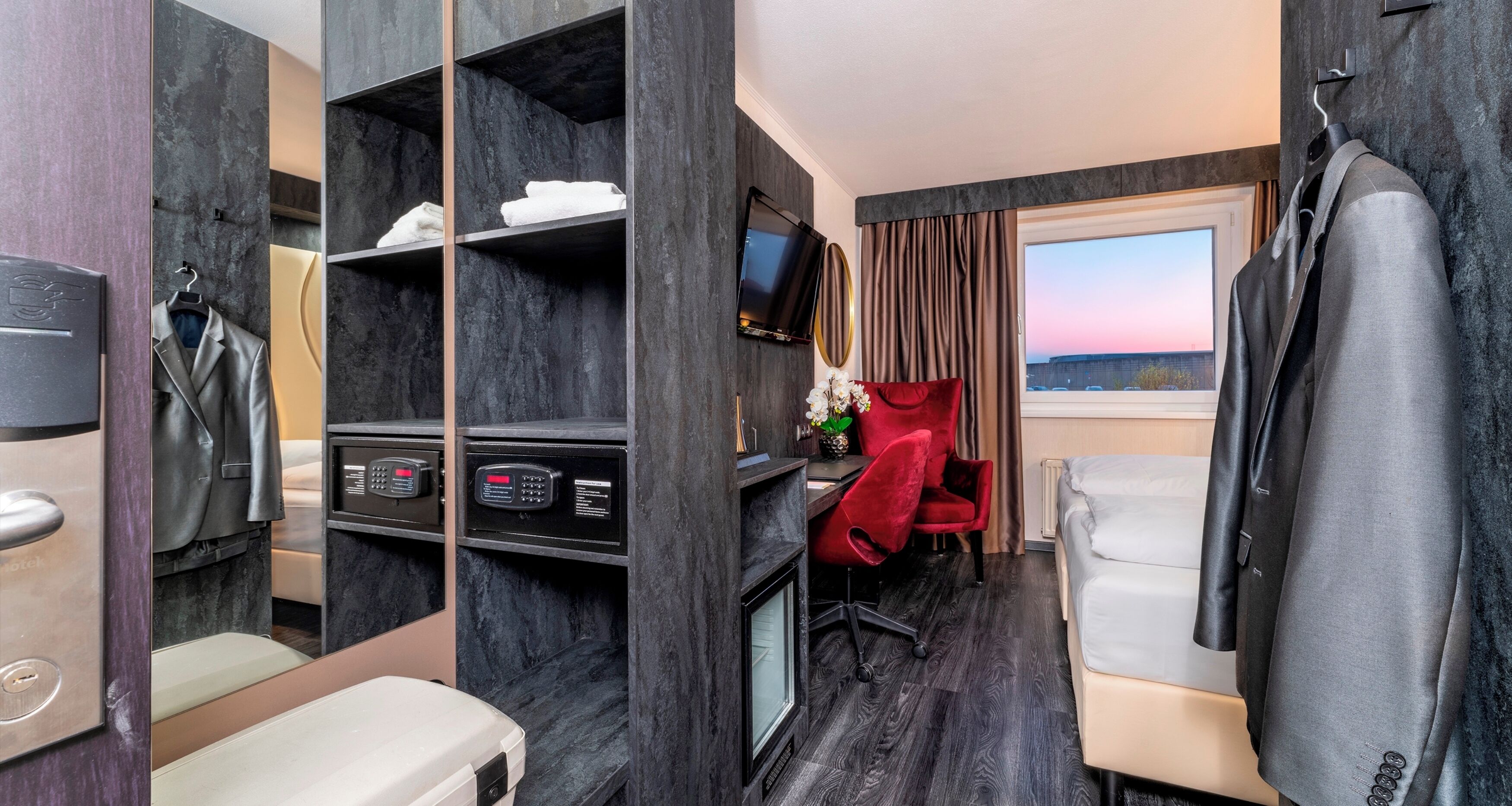 Quarto de hotel com uma cama, uma escrivaninha com um computador, uma TV, uma cadeira vermelha, um sofá vermelho e vista do céu.