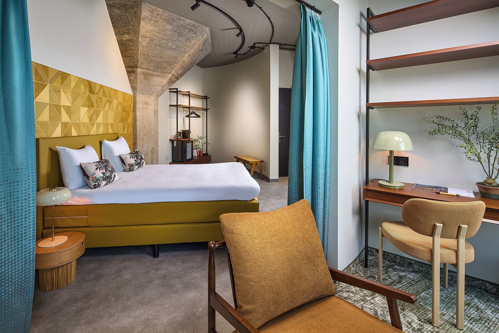 Quarto de hotel moderno com uma cama, uma escrivaninha, uma cadeira, um abajur e um vaso de plantas.