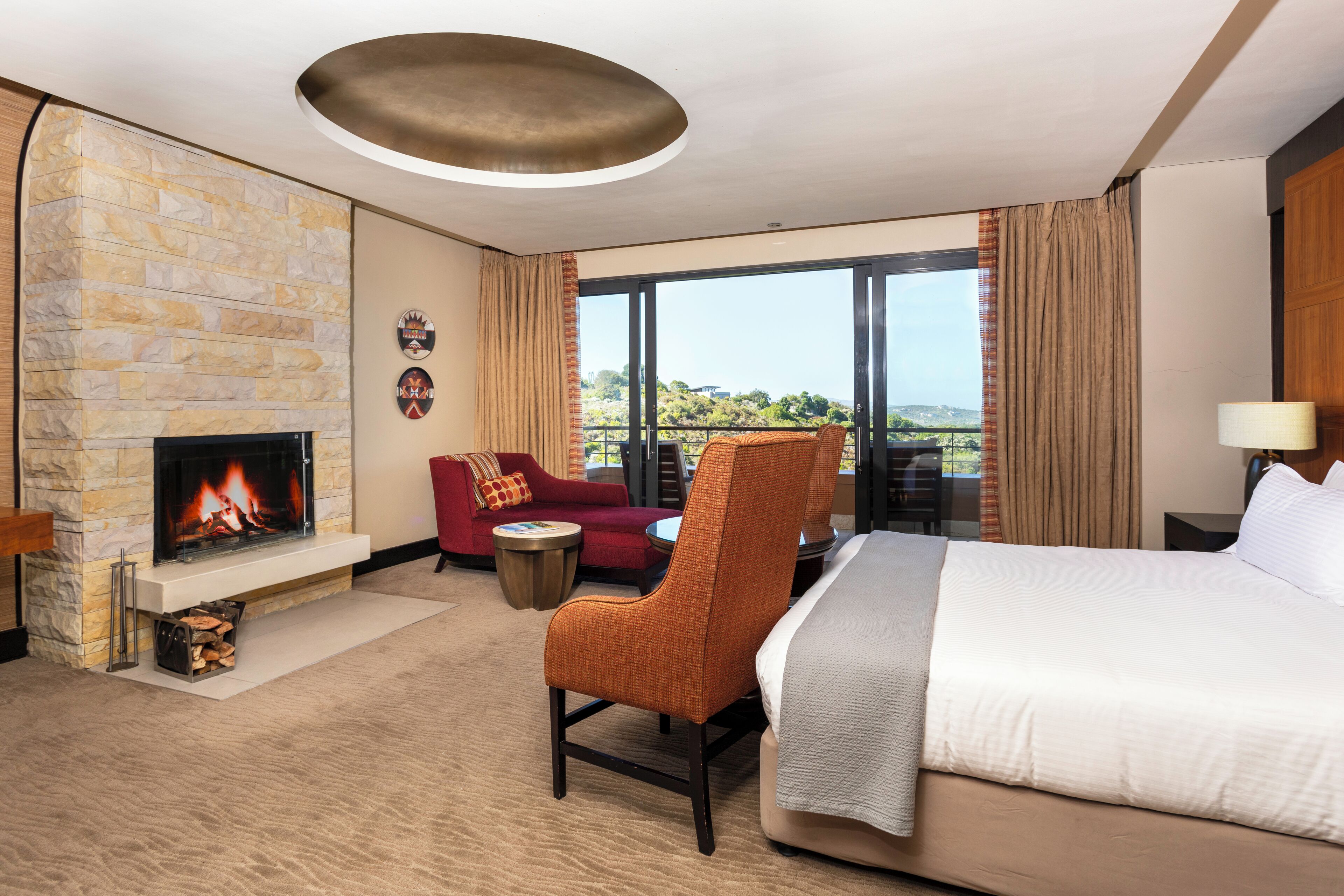 Quarto de hotel com lareira de pedra, uma cama grande, uma poltrona vermelha e vista para o exterior.