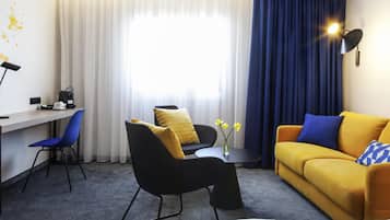 Quarto de hotel com escrivaninha, cadeira, sofá, cama, luminária e janela com cortinas.