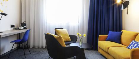 Quarto de hotel com escrivaninha, cadeira, sofá, cama, luminária e janela com cortinas.