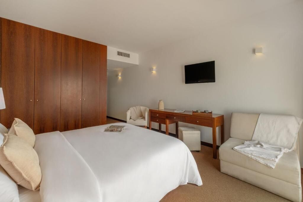Quarto de hotel com uma cama grande, uma escrivaninha, uma cadeira, um sofá e uma TV fixada na parede.