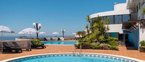 Uma área de piscina com espreguiçadeiras e guarda-sóis, vista desobstruída para o oceano e um edifício moderno com grandes janelas.