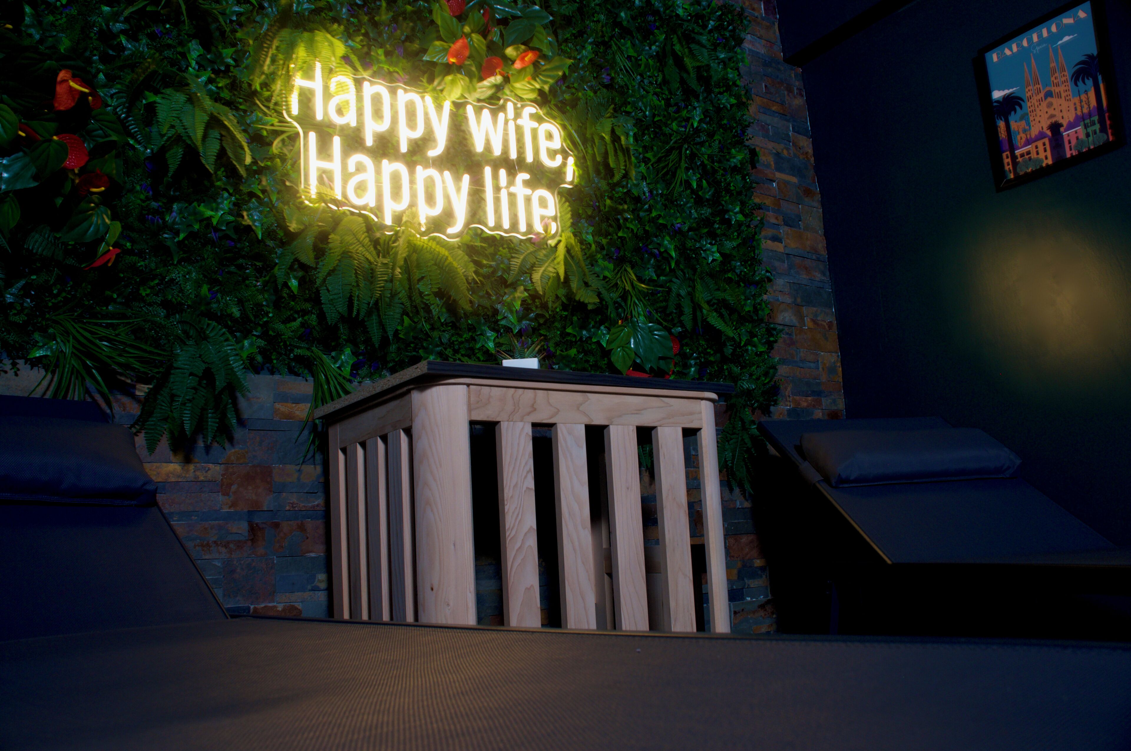 Quarto com um letreiro de neon que diz 'Happy wife, Happy life!' acima de um banco de madeira.
