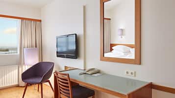 Quarto de hotel com escrivaninha, cadeira, televisão e um espelho grande que reflete a cama.
