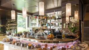 Uma área de bar com balcão de mármore, diversos alimentos e bebidas, e uma estante bem abastecida com garrafas e copos.