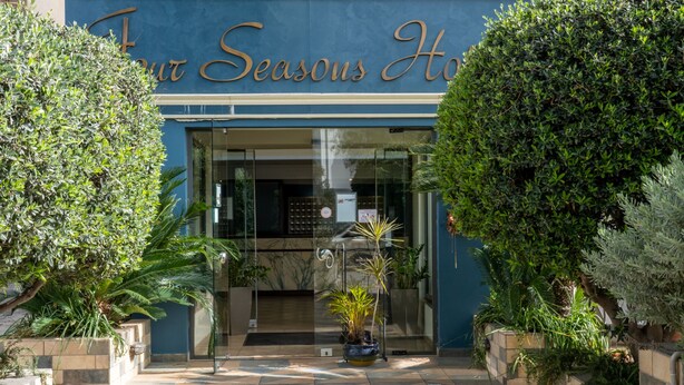 Entrada de um hotel com fachada azul e a inscrição 'Four Seasons Hotel' em dourado. Há plantas em vasos e um caminho de ladrilhos levando à entrada.