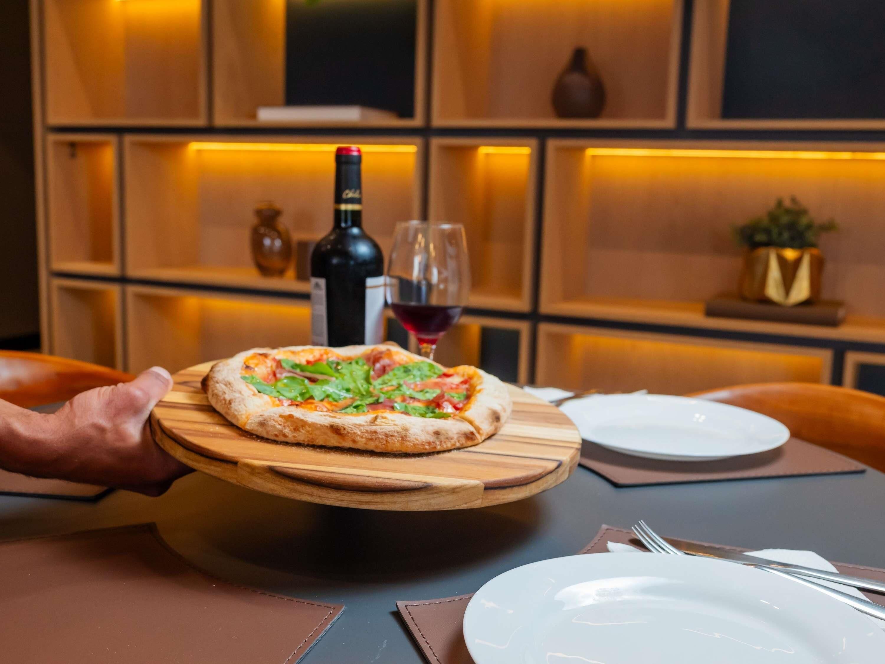Pizza em tábua de madeira, uma garrafa de vinho e um copo de vinho tinto sobre a mesa.
