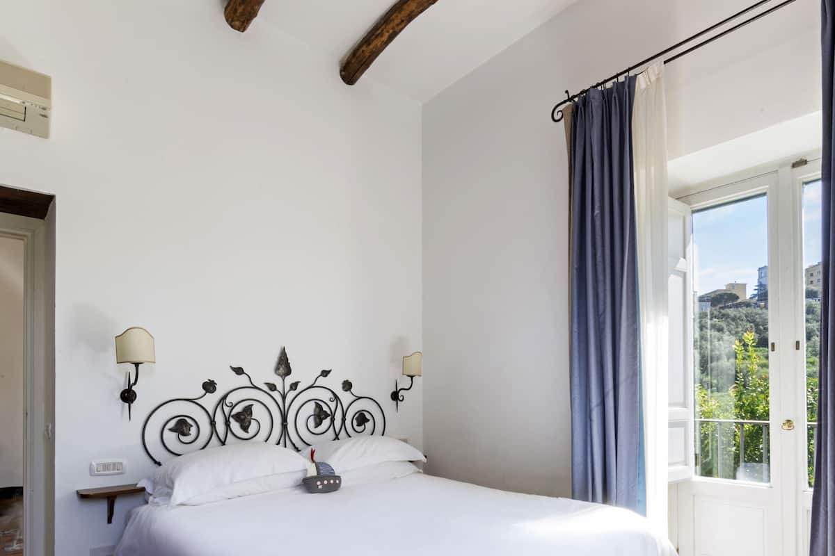 Quarto com cama, mesa de cabeceira, luminária na parede e porta que dá para uma varanda.