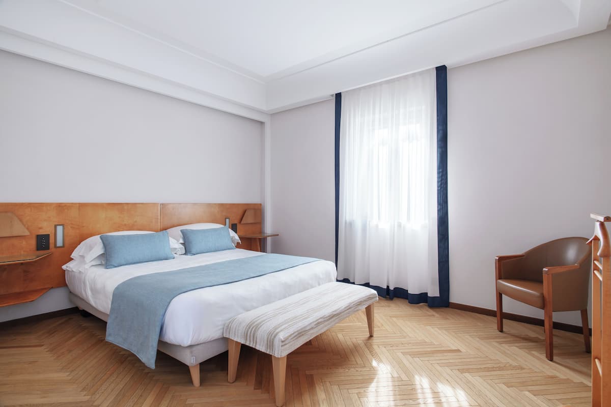Quarto de hotel com cama de cabeceira de madeira, um banco, uma cadeira de madeira e uma mesinha de cabeceira com abajur.