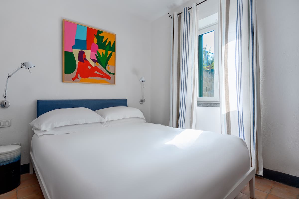 Uma cama bem arrumada com lençóis brancos, cabeceira azul e um quadro abstrato colorido na parede.