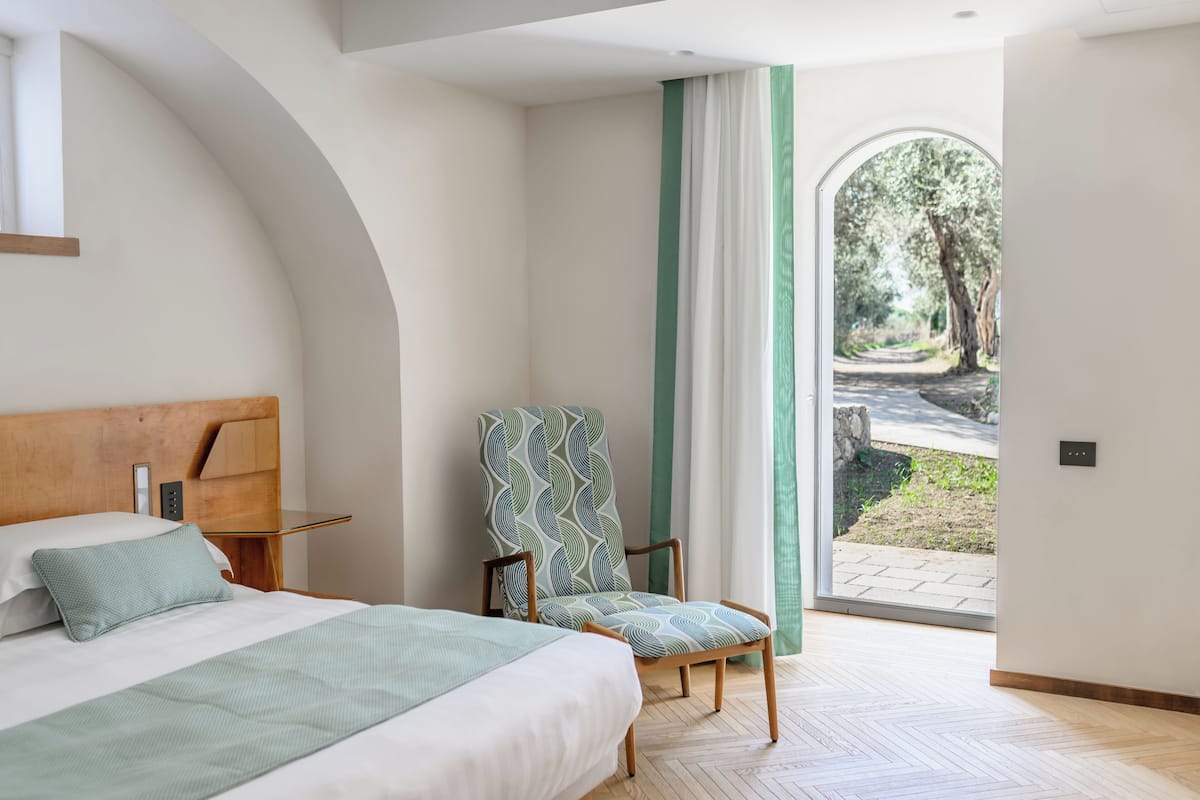 Quarto com cama, uma cadeira e vista para um jardim pela janela.