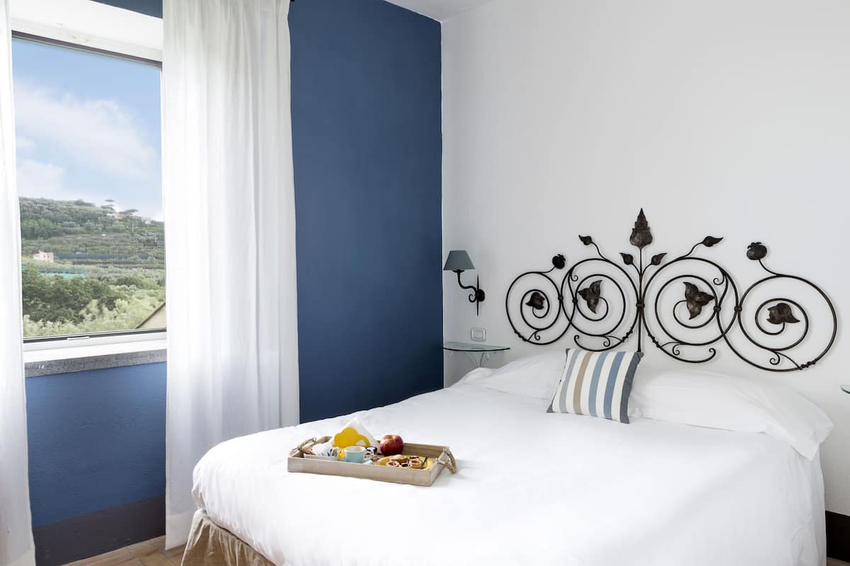 Quarto com cama, bandeja de itens de café da manhã, janela com cortinas e vista para o verde lá fora.