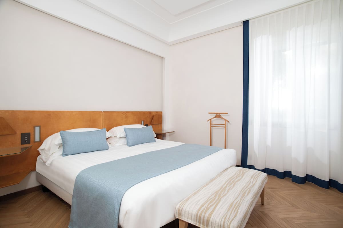 Quarto de hotel com cama grande, mesa de cabeceira, cabideiro e janela com cortinas leves.