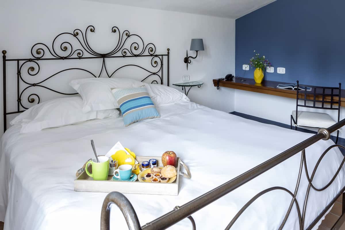 Cama bem arrumada com lençóis brancos, bandeja de itens de café da manhã incluindo torradas, frutas e uma xícara, e uma mesa de cabeceira com abajur e um vaso de flores.