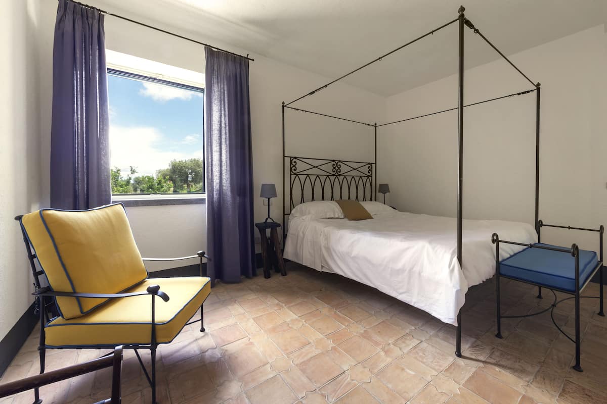 Quarto com cama de dossel, uma cadeira amarela, uma mesinha e uma janela com cortinas.