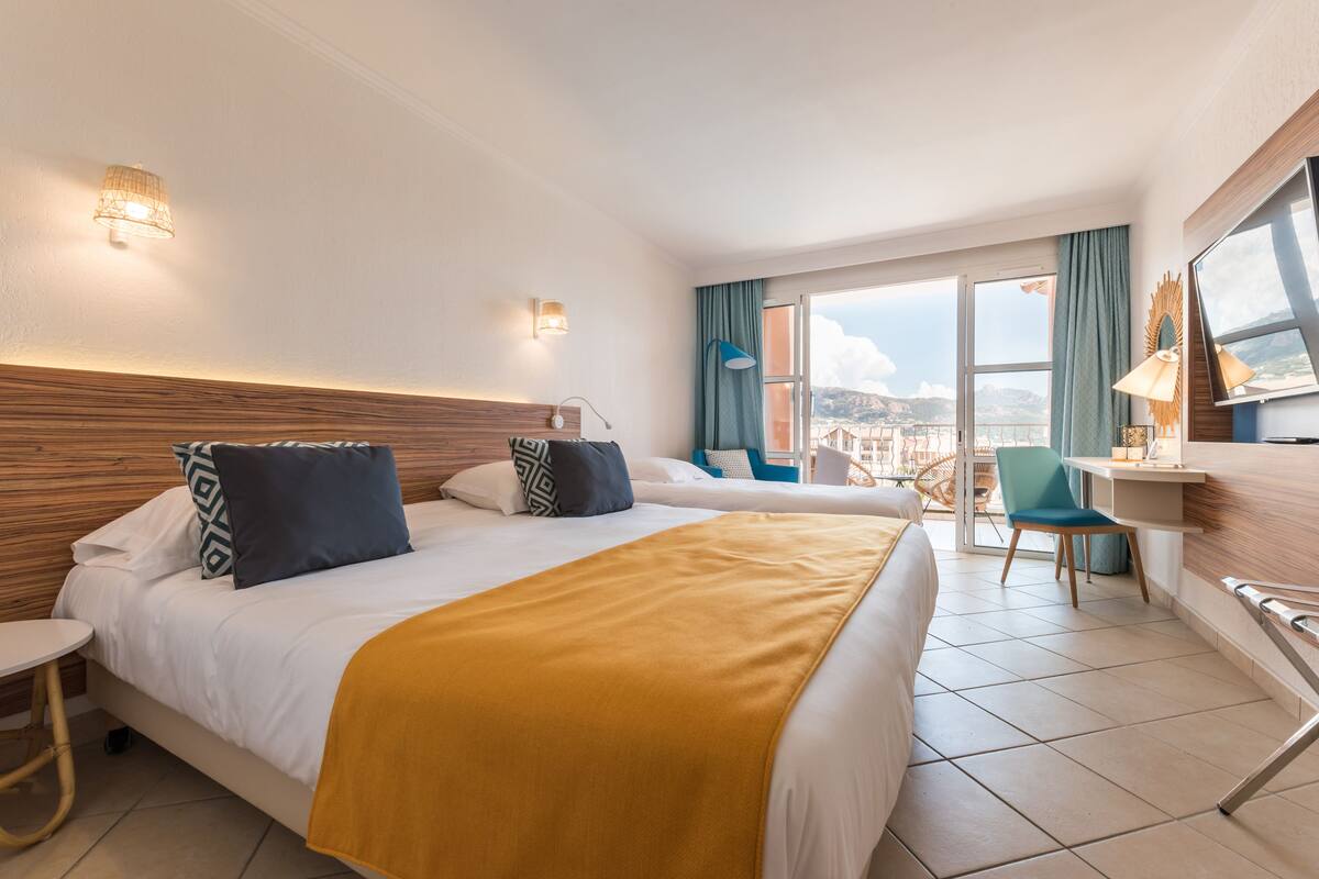 Quarto de hotel com duas camas, uma escrivaninha, uma cadeira e uma janela grande com vista.