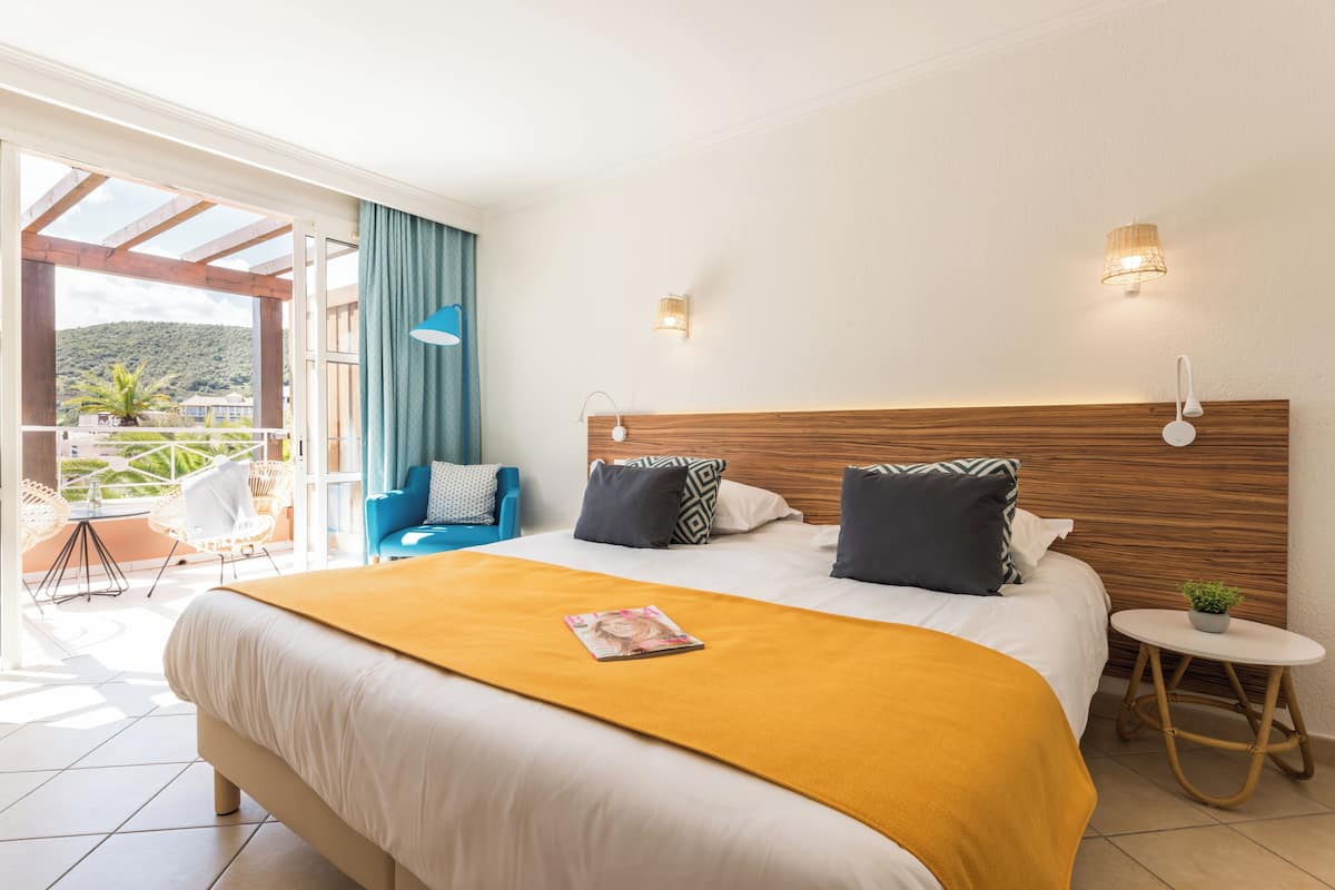 Quarto de hotel com cama, uma pequena mesa redonda com uma planta, varanda com cadeiras e vista para o verde.