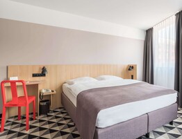 Quarto de hotel com cama, escrivaninha, cadeira, luminária e lixeira.
