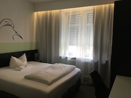 Uma cama bem arrumada com almofadas e um cobertor dobrado, uma mesa de cabeceira com abajur, e uma janela com cortinas leves.