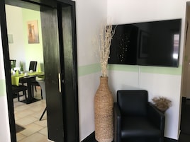 Uma área de recepção com uma porta preta, um vaso trançado com galhos, uma cadeira preta e uma televisão de tela plana na parede.