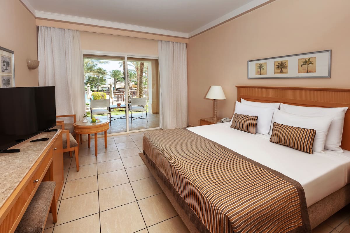 Quarto de hotel com cama, escrivaninha, televisão, poltrona e vista para a área externa.