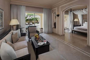Quarto de hotel com sofá, mesa de centro com frutas, cama com dossel e vista para área externa com guarda-sóis.