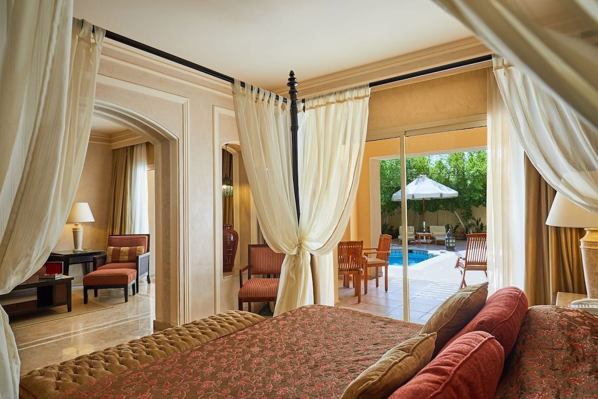 Quarto de hotel com cama, poltrona, mesa, abajur e vista para uma piscina ao ar livre.