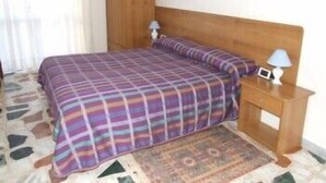 Quarto com cabeceira de madeira, cama com colcha roxa xadrez, duas luminárias de mesa e um tapete estampado.