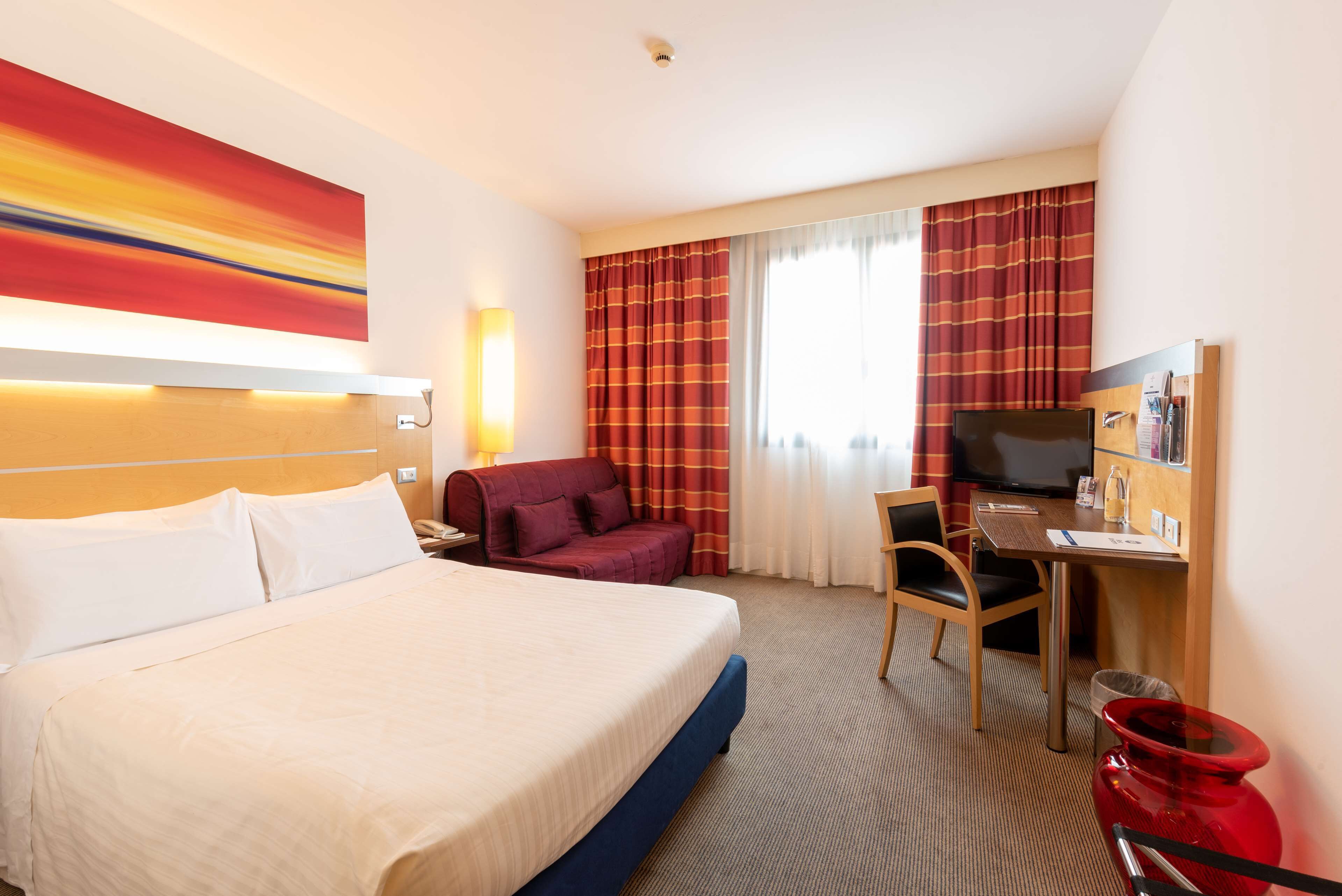 Quarto de hotel com uma cama grande, uma escrivaninha com cadeira, televisão e uma lixeira vermelha.