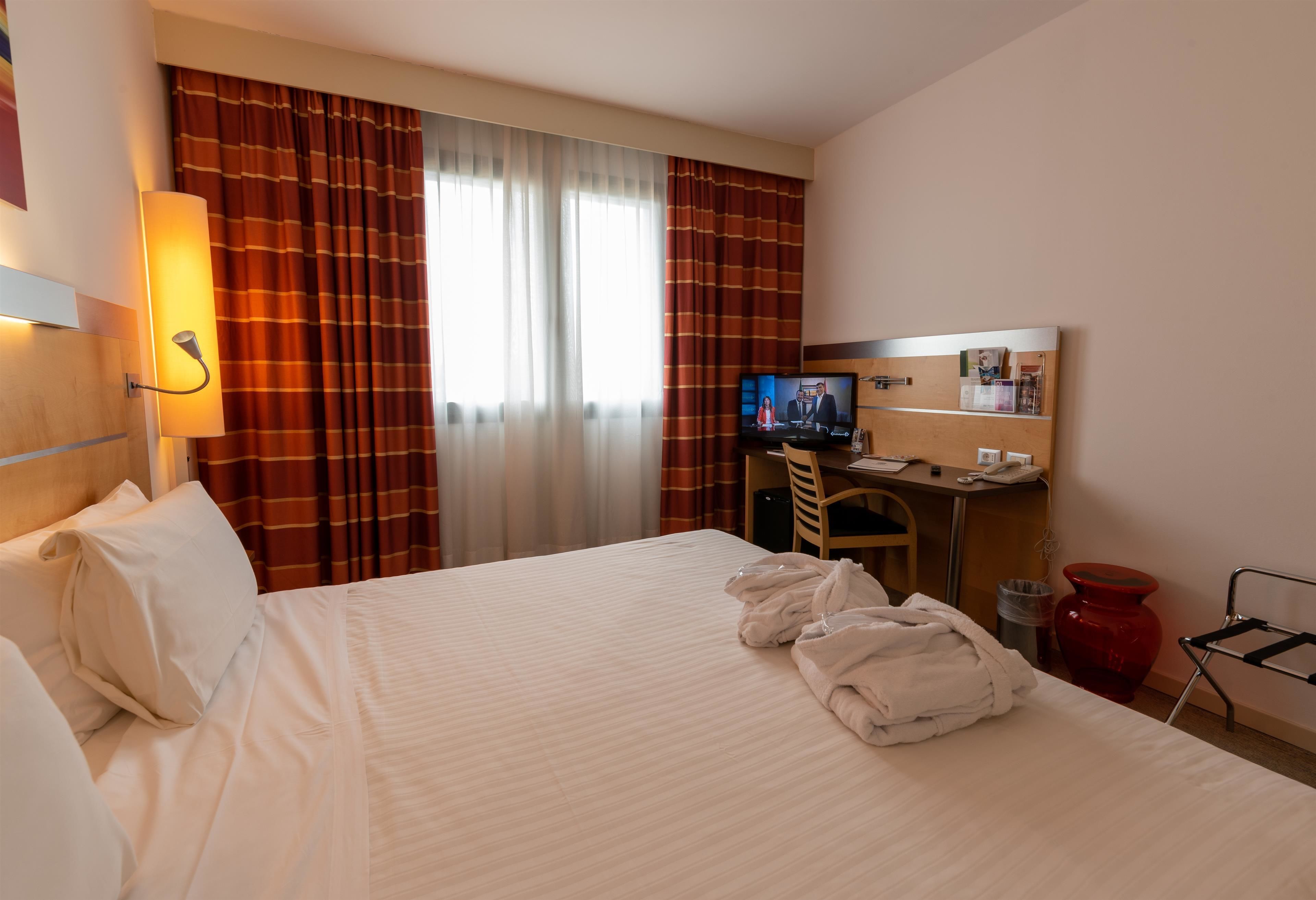Quarto de hotel com cama, escrivaninha, televisão e janela com cortinas.