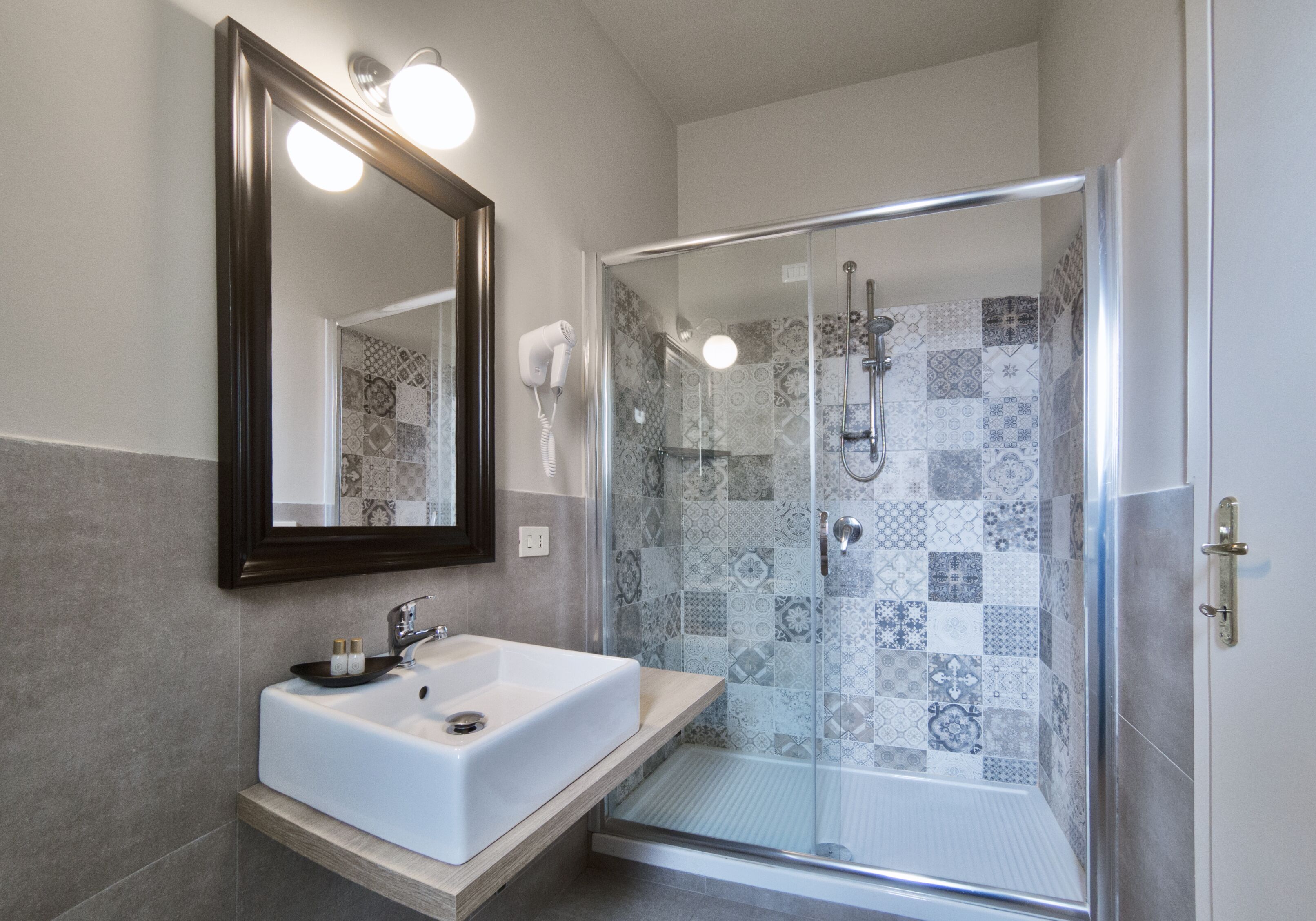 Casa de banho com lavatório branco, espelho e chuveiro com azulejos estampados.