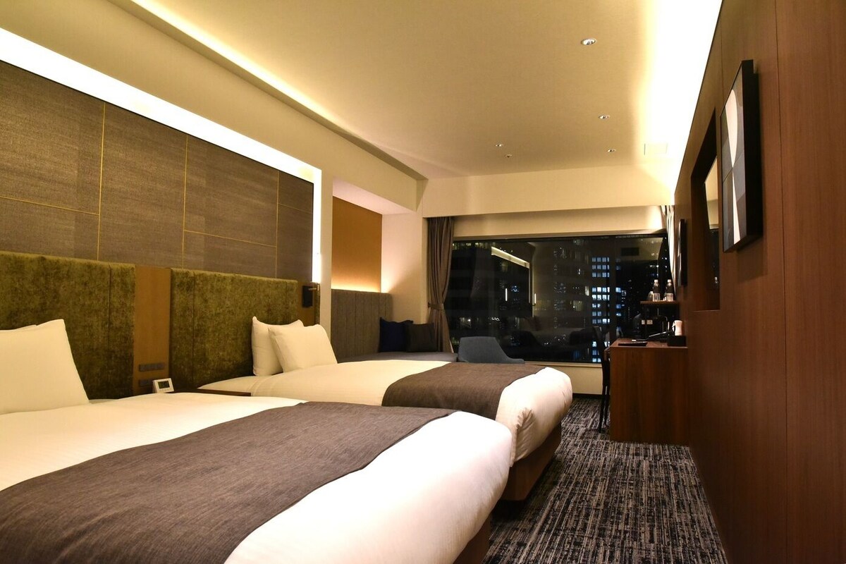 Quarto de hotel com duas camas, vista para a cidade e escrivaninha com cadeira.