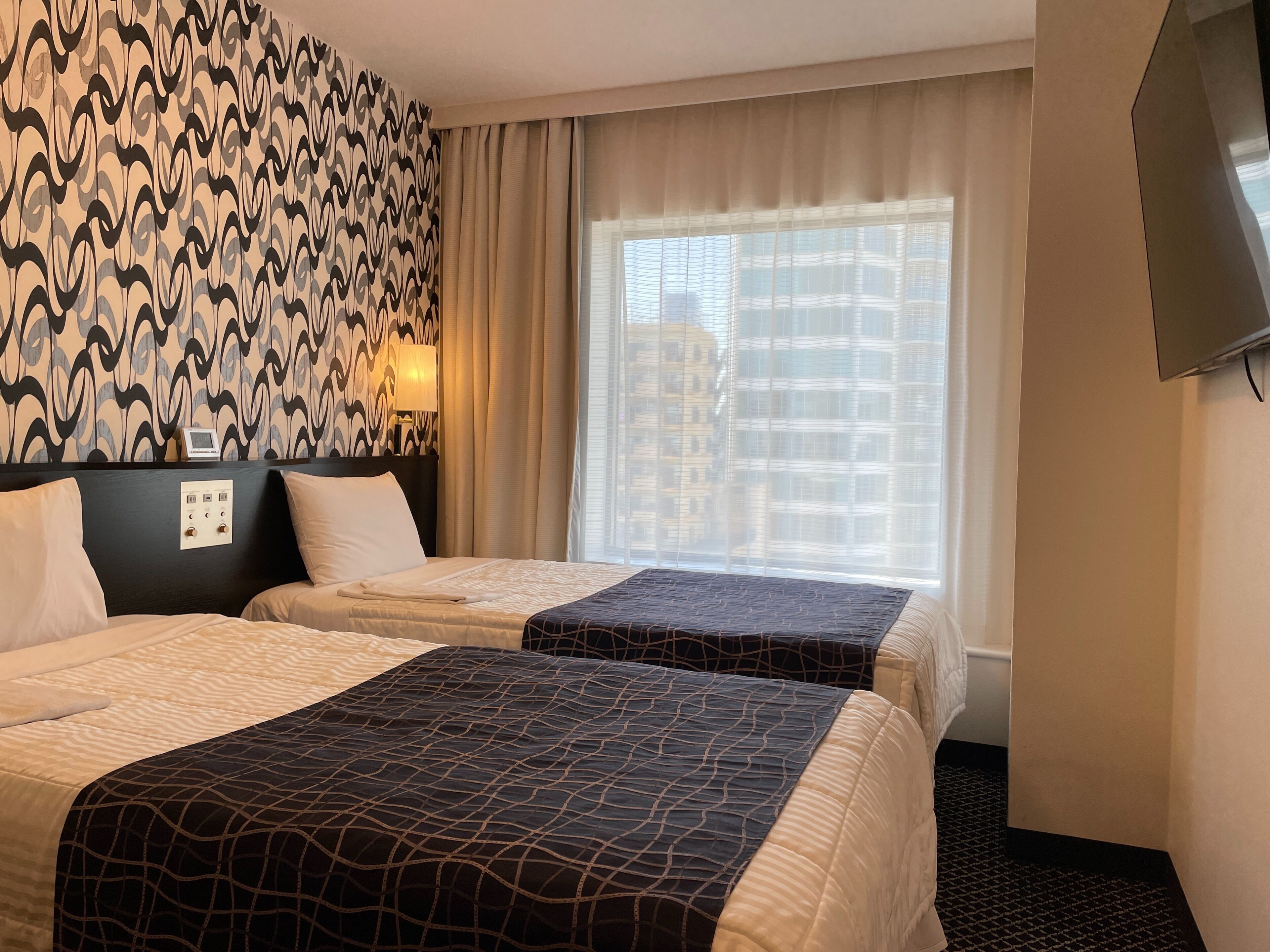 Quarto de hotel com duas camas, parede estampada, janela grande com cortinas e televisão de tela plana fixada na parede.